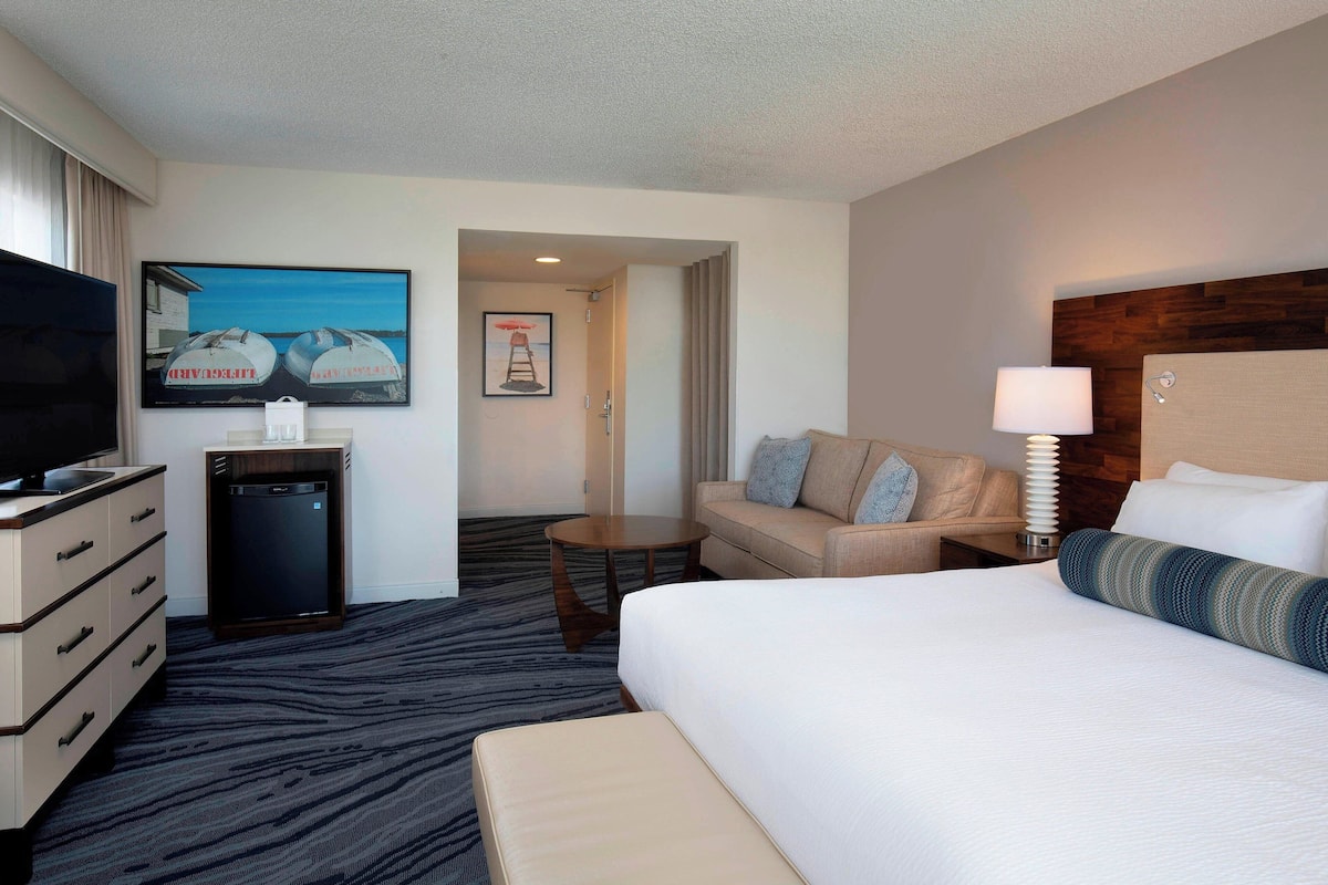 Quarto de hotel com cama, televisão, secretária com luminária, sofá e um quadro na parede.