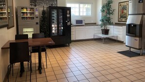 Uma cozinha moderna com armários brancos, uma área de jantar com mesa e cadeiras, micro-ondas, geladeira, máquina de vendas e uma planta em vaso.