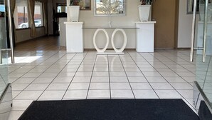 Uma área de recepção com piso de azulejos, duas plantas em vasos, um balcão de recepção e um espelho grande na parede.