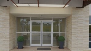 Entrada de um edifício com porta de vidro, duas plantas em vasos e estrutura de telhado em metal.