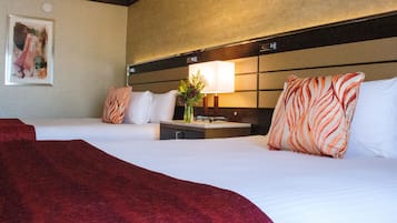 Quarto de hotel com duas camas, uma mesinha de cabeceira, uma luminária e um quadro na parede.