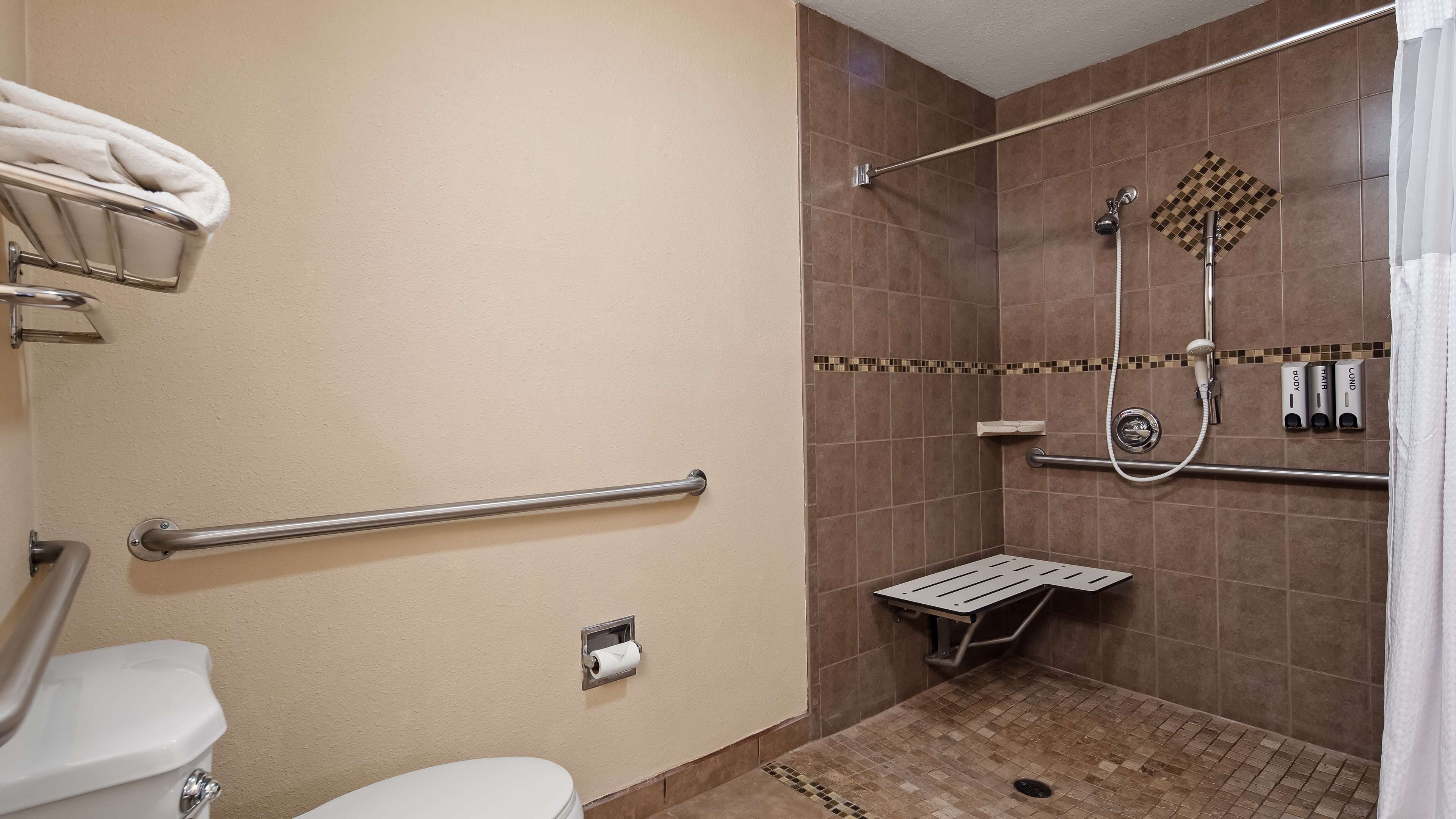Casa de banho adaptada para pessoas com deficiência, com sanita, barras de apoio, chuveiro com esguicho manual e um porta-toalhas.