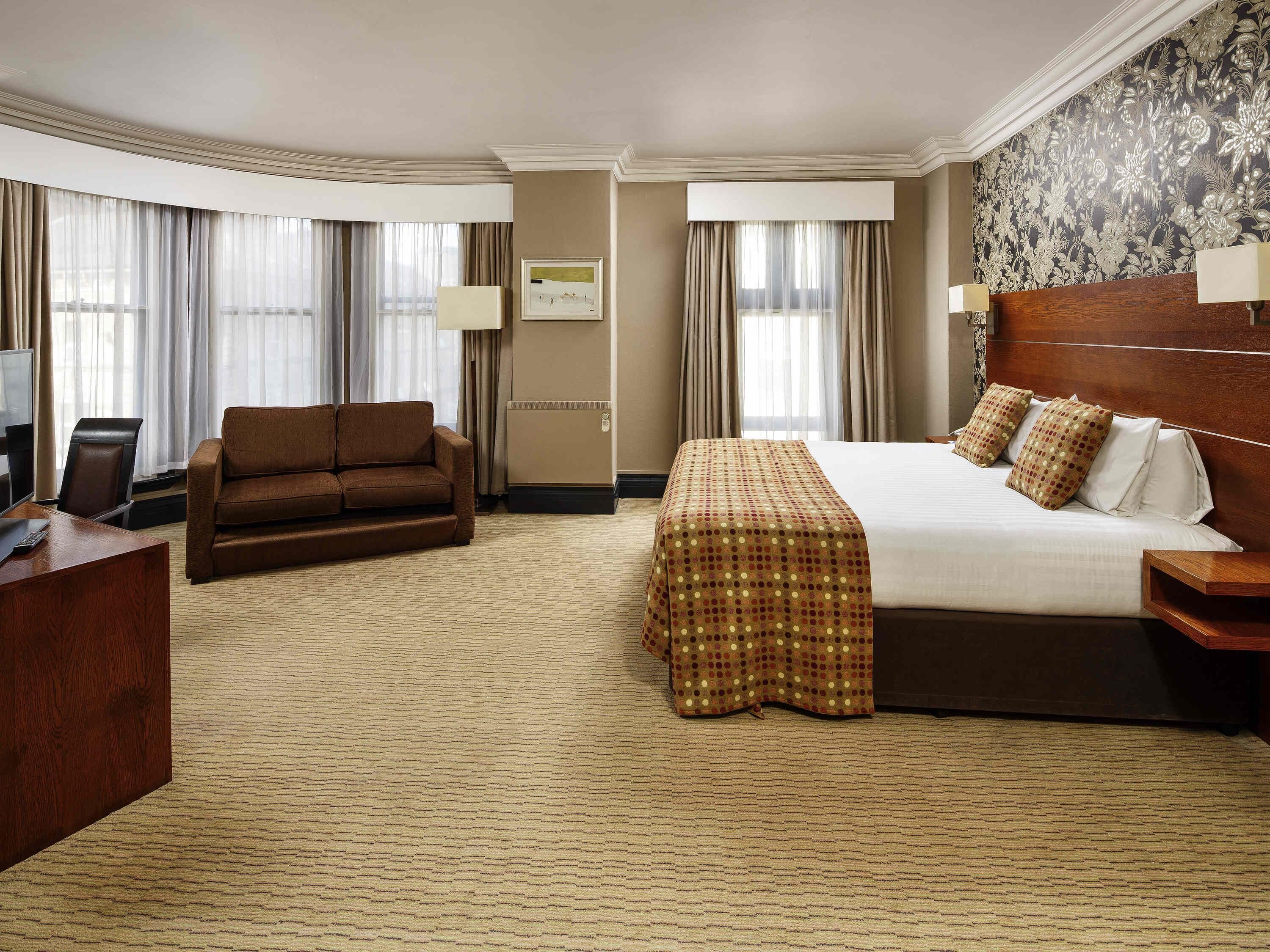 Quarto de hotel com cama, sofá, escrivaninha e televisão.