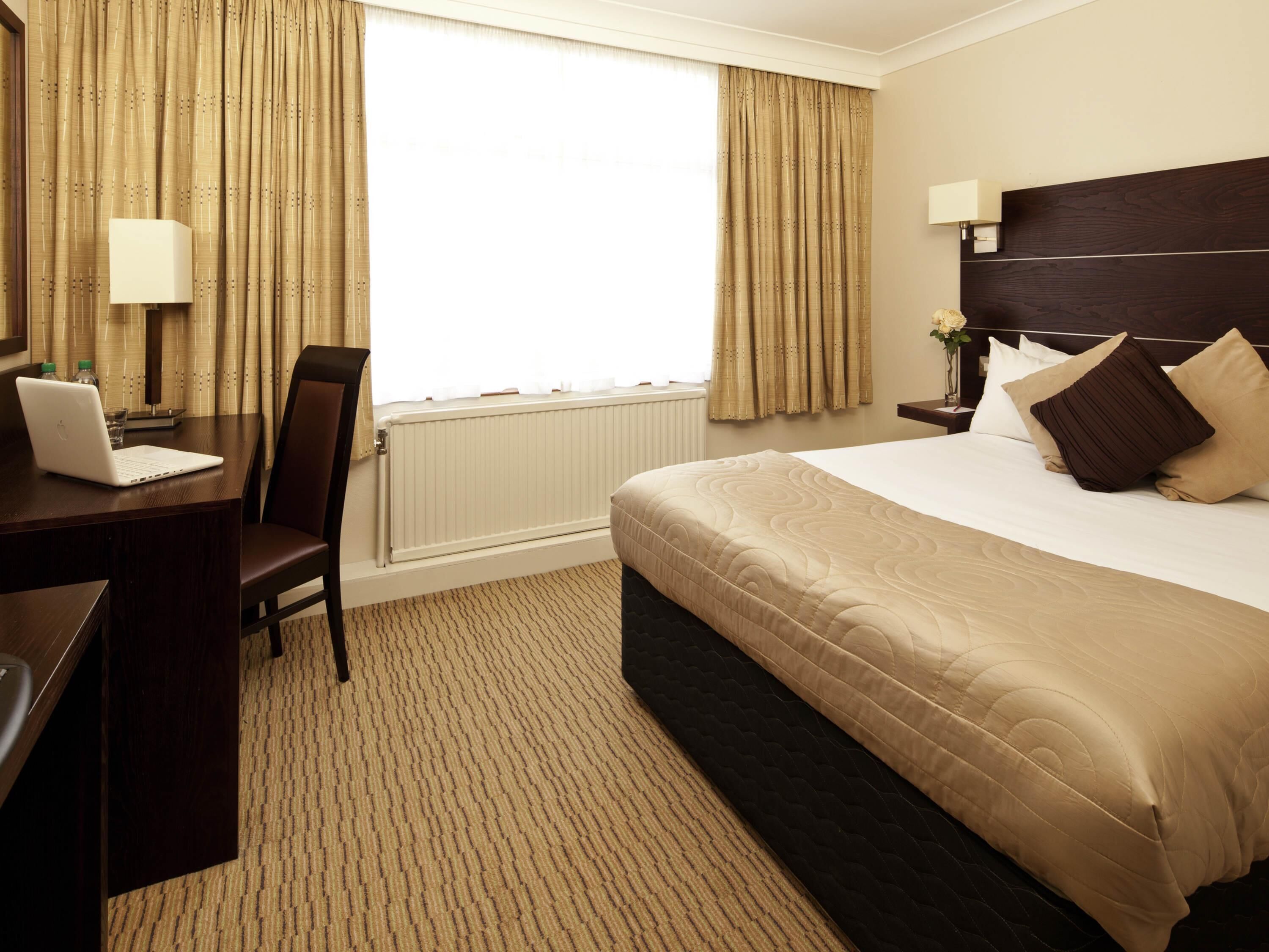 Quarto de hotel com cama, escrivaninha com laptop, cadeira, mesa de cabeceira com luminária e janela com cortinas.