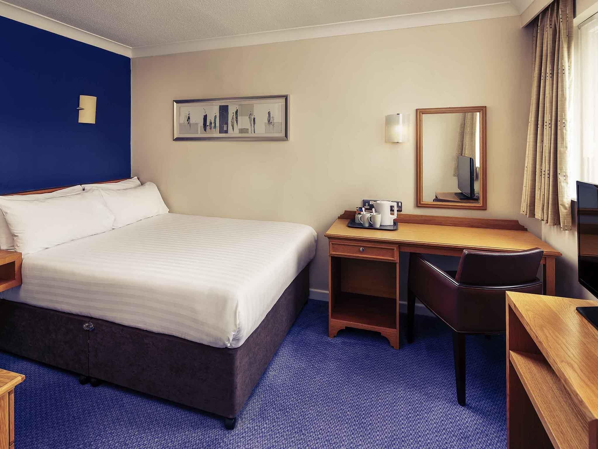 Quarto de hotel com cama, escrivaninha, cadeira, máquina de café, televisão e quadro na parede.