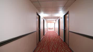 Corredor de hotel com carpete estampado, portas em ambos os lados e teto com iluminação embutida.
