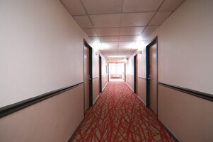 Corredor de hotel com carpete estampado, portas em ambos os lados e teto com iluminação embutida.