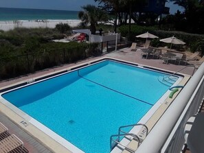 Uma piscina retangular com água azul cristalina, rodeada por espreguiçadeiras e guarda-sóis, com vista para a praia e para o oceano ao fundo.