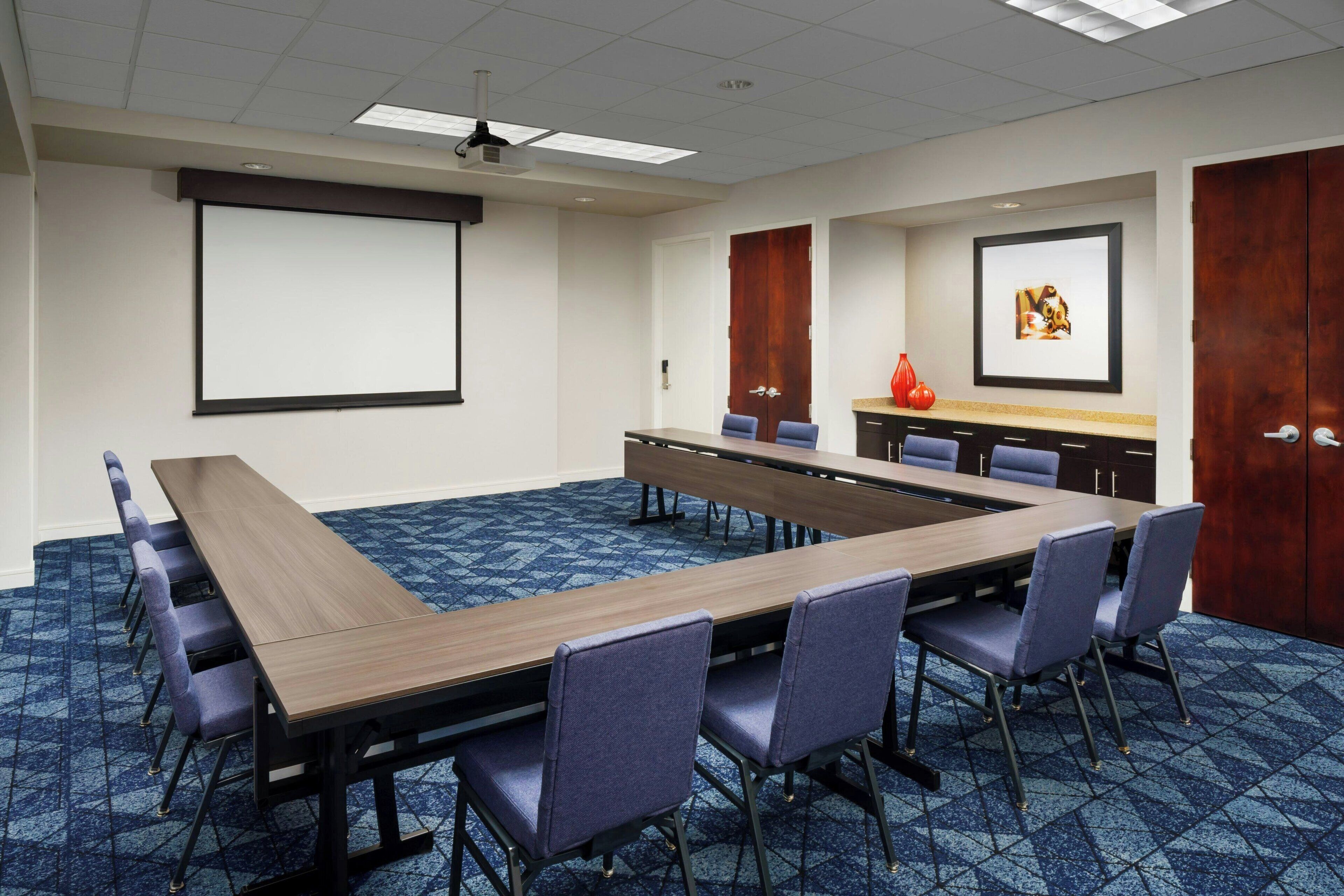 Sala de conferência com uma mesa comprida, carpete estampado em azul e uma tela de projetor.