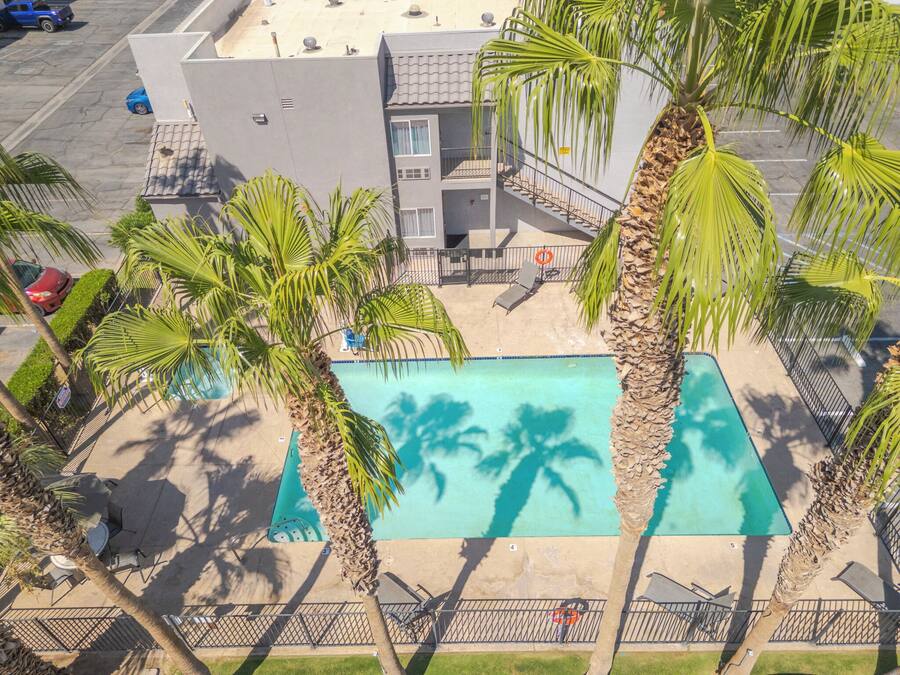 Piscina no terraço com palmeiras, um edifício com fachada cinzenta e área de estacionamento com carros.