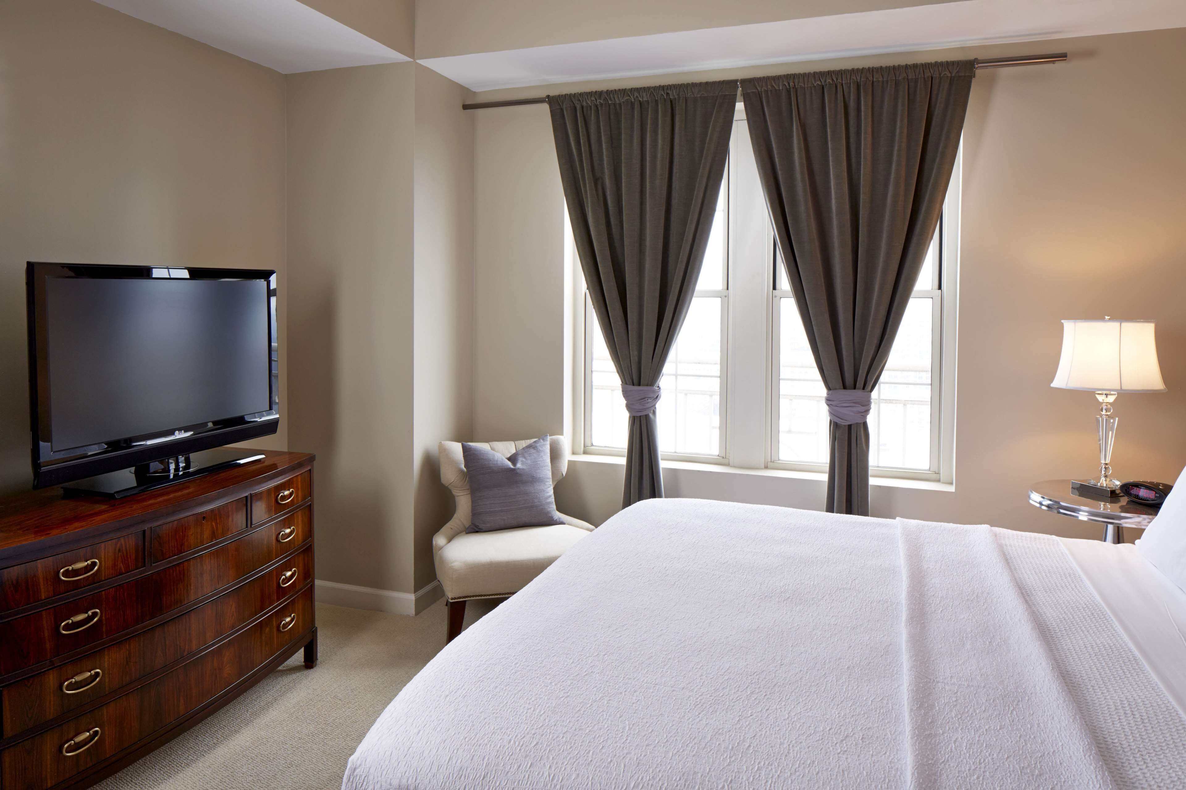 Quarto de hotel com televisão de ecrã plano sobre uma cómoda de madeira, uma cama com roupa de cama branca, uma cadeira com almofada e um candeeiro num apoio de mesa.