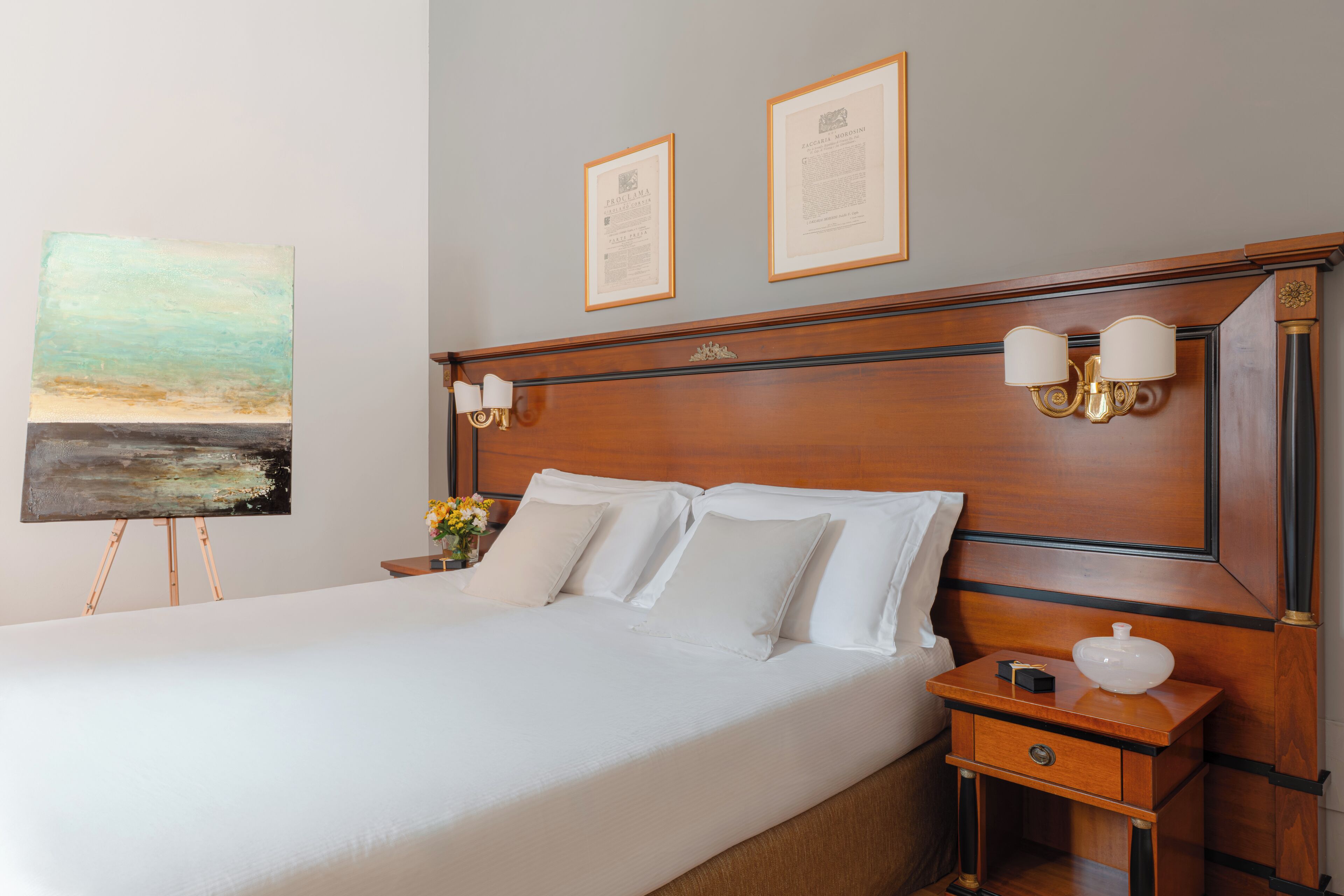 Cama bem arrumada com lençóis brancos, cabeceira de madeira, dois certificados emoldurados na parede e mesa de cabeceira com abajur e um pequeno vaso de flores.