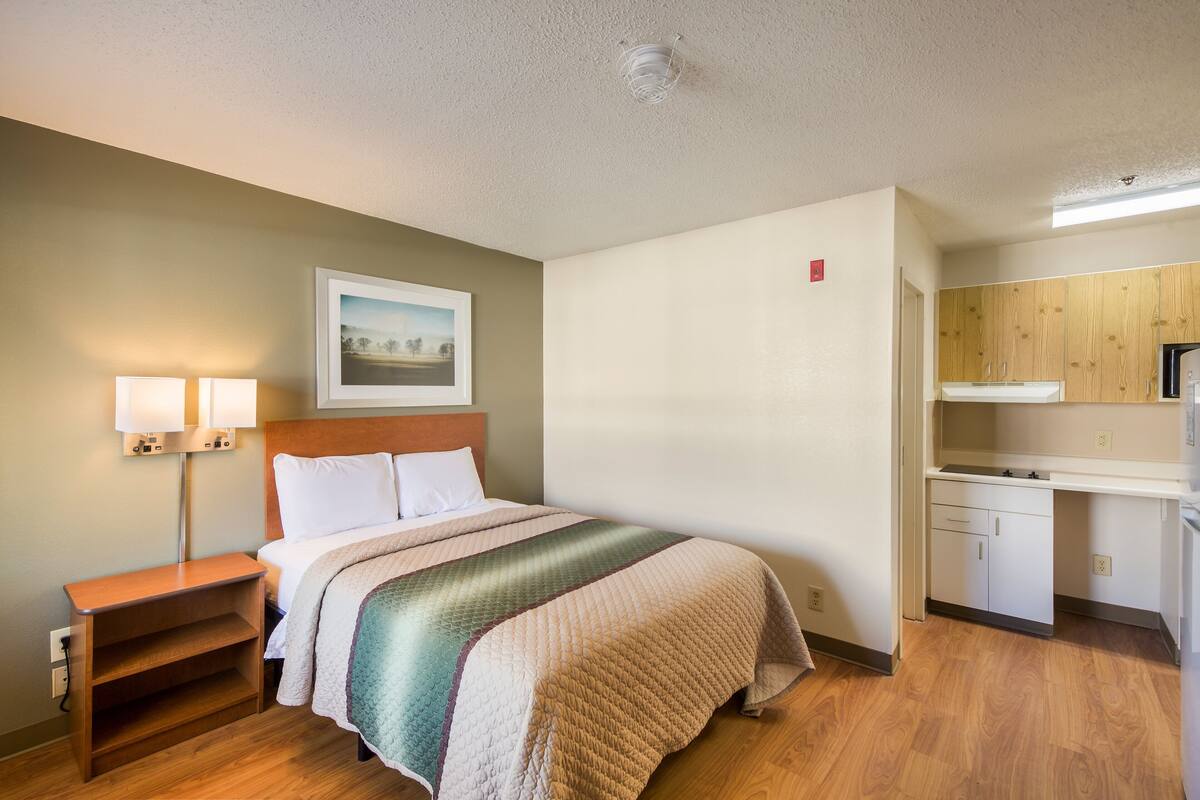Quarto de hotel com cama, luminárias de cabeceira, mesa de apoio, pintura na parede e uma pequena cozinha.