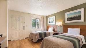 Quarto de hotel com duas camas, uma escrivaninha, uma cadeira, janela com cortinas, porta e um quadro emoldurado na parede.
