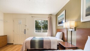 Quarto de hotel com duas camas, uma janela com cortinas, uma mesa de cabeceira com abajur e uma porta com aviso.