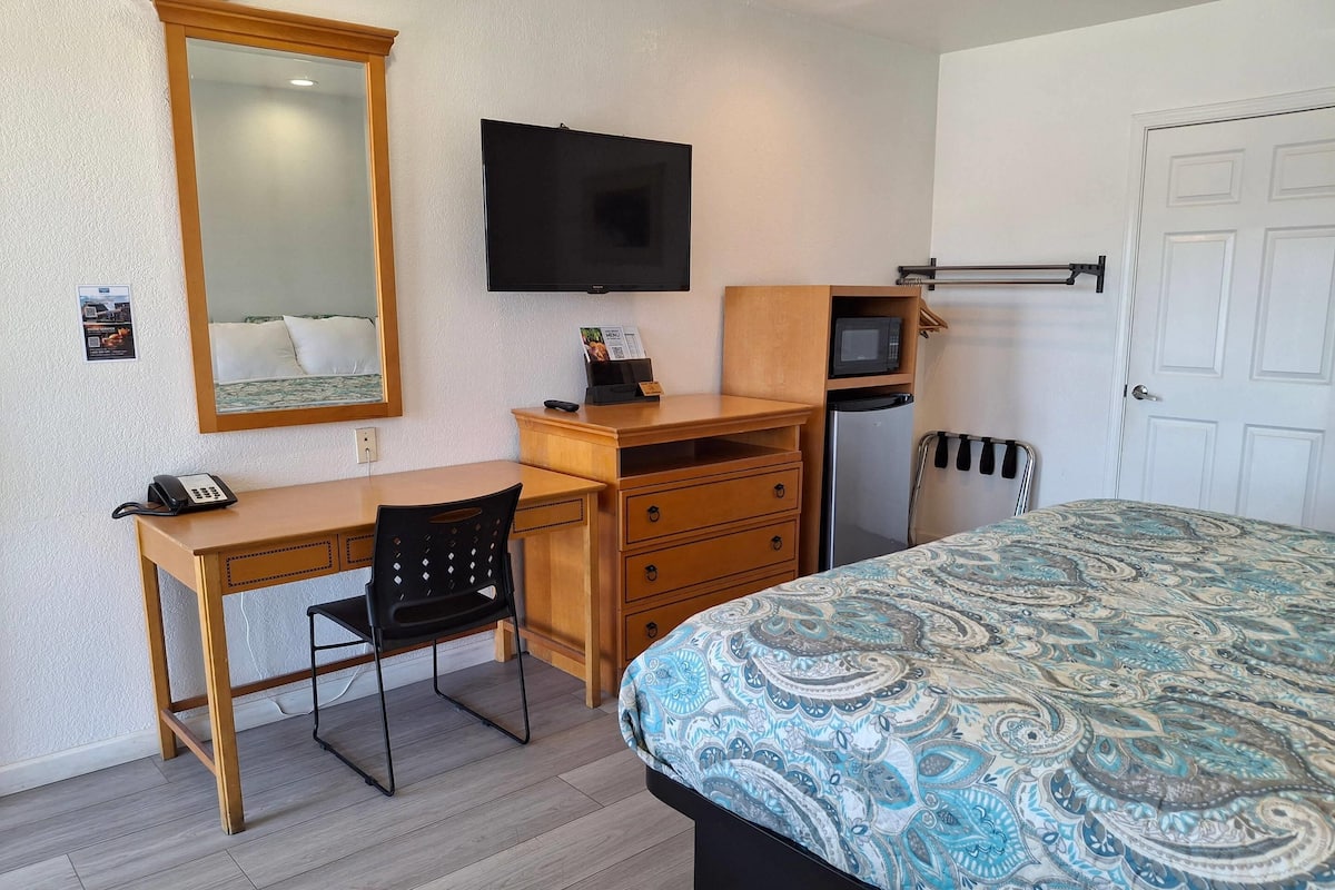 Quarto de hotel com escrivaninha de madeira, uma cadeira, cama com colcha estampada, televisão, micro-ondas, frigorífico e porta.