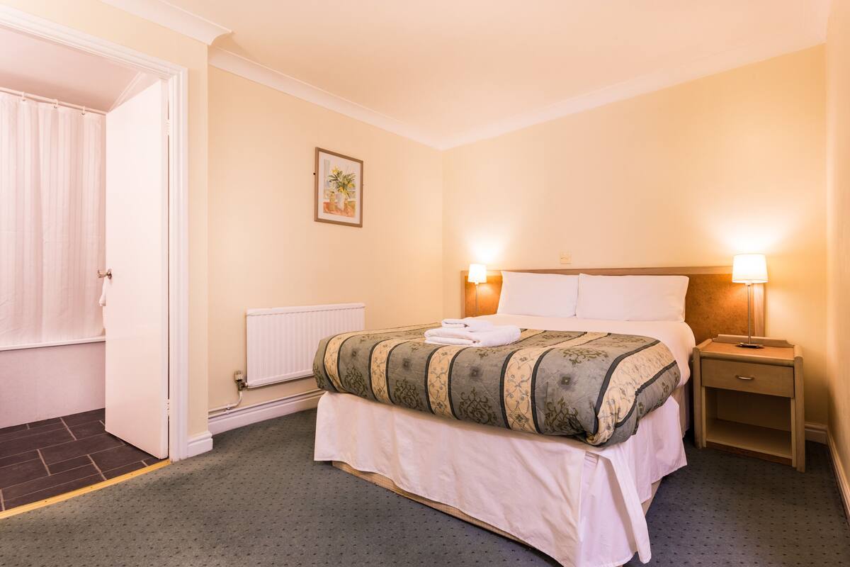 Quarto de hotel com cama, abajures, radiador e porta.