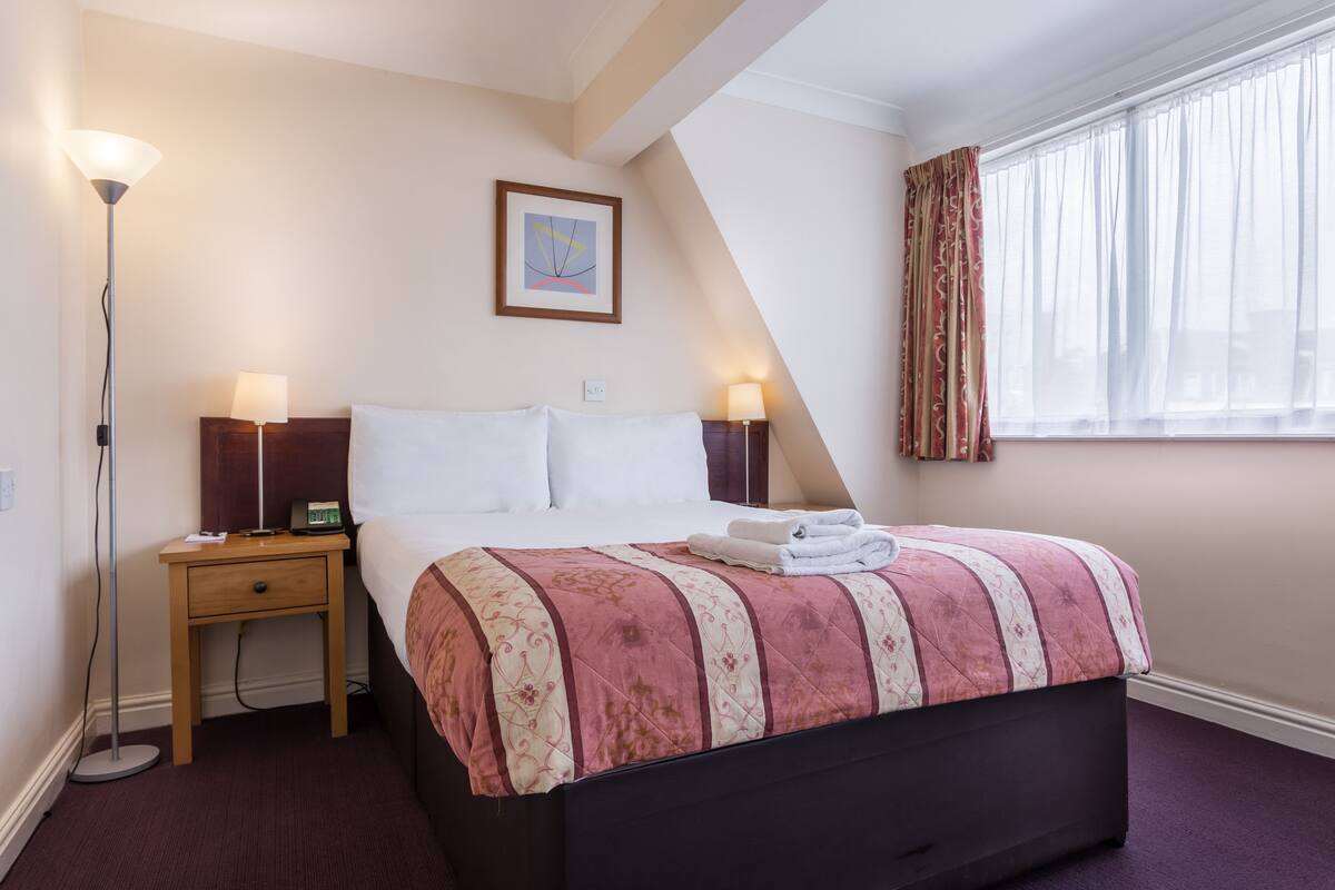 Quarto de hotel com cama, luminárias laterais, mesa de cabeceira, quadro na parede e janela com cortinas.