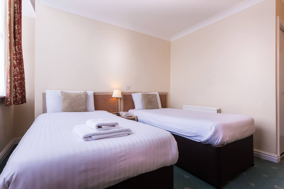 Quarto de hotel com duas camas, uma mesa de cabeceira com abajur e uma janela com cortinas.