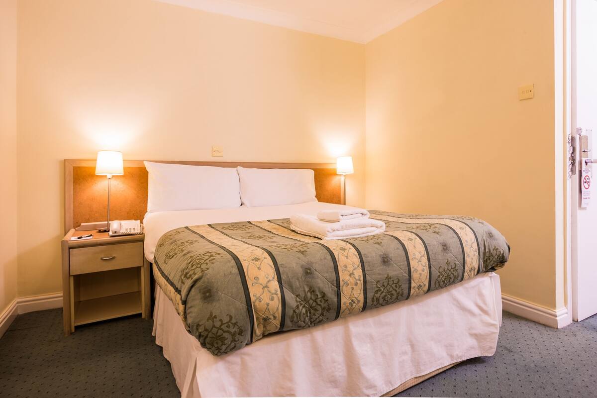 Quarto de hotel com cama, duas luminárias de cabeceira, mesa de apoio e porta.