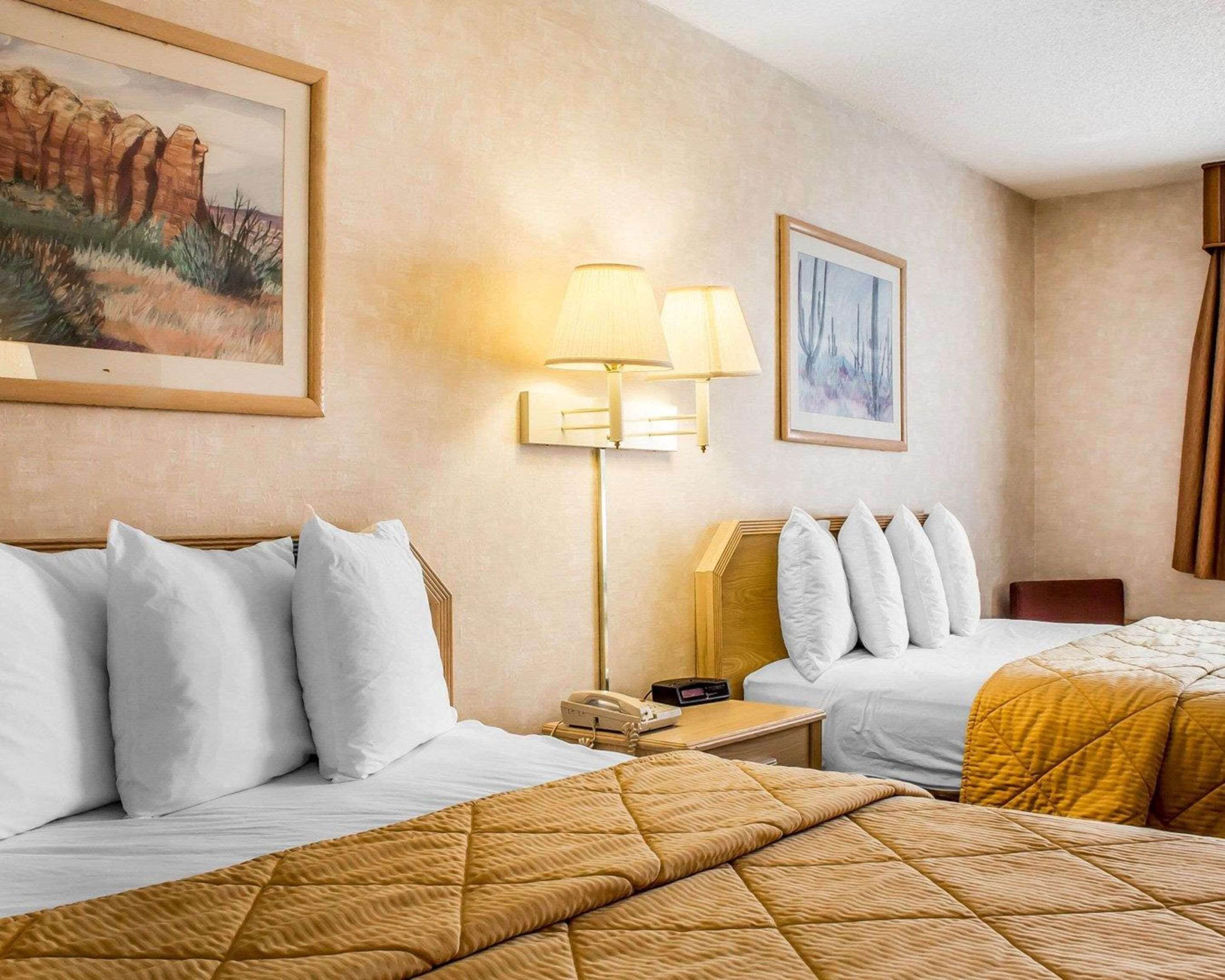 Quarto de hotel com duas camas, uma mesinha de cabeceira com abajur, um quadro na parede e uma janela com cortinas.