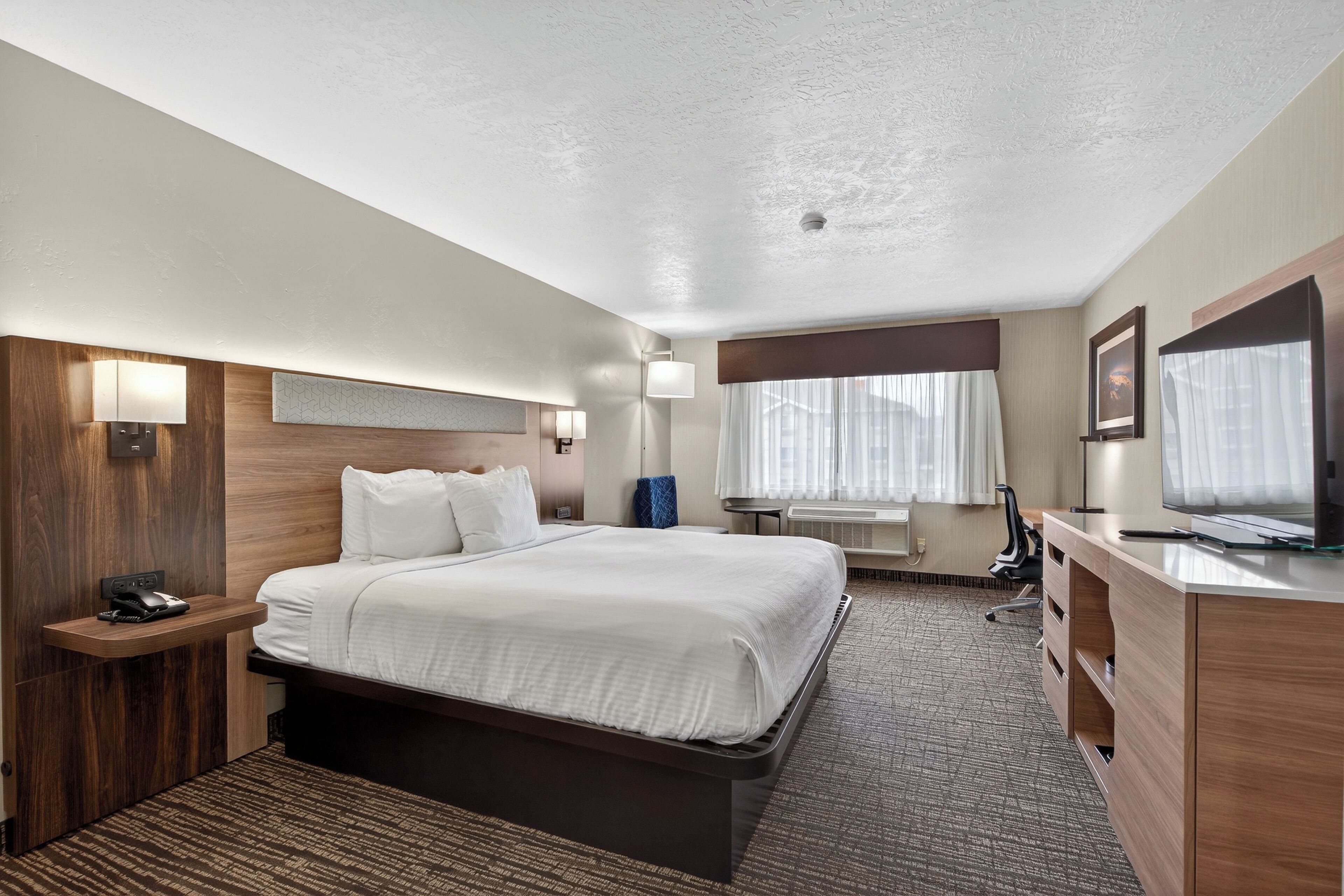 Quarto de hotel com uma cama grande, uma escrivaninha, uma cadeira, uma televisão e uma janela com cortinas.