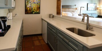 Cozinha moderna com pia de aço inoxidável, bancadas brancas e vista para um quarto através de uma janela.