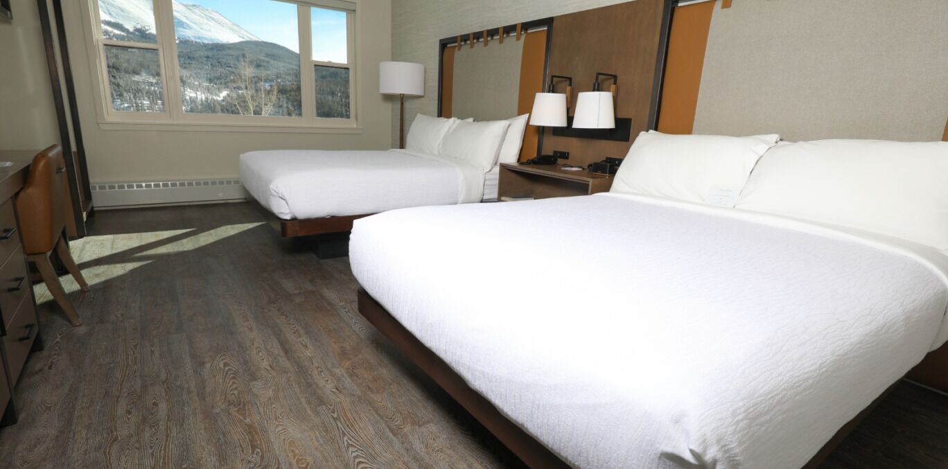 Quarto de hotel com duas camas, uma escrivaninha, uma cadeira, um abajur e uma janela com vista para as montanhas.