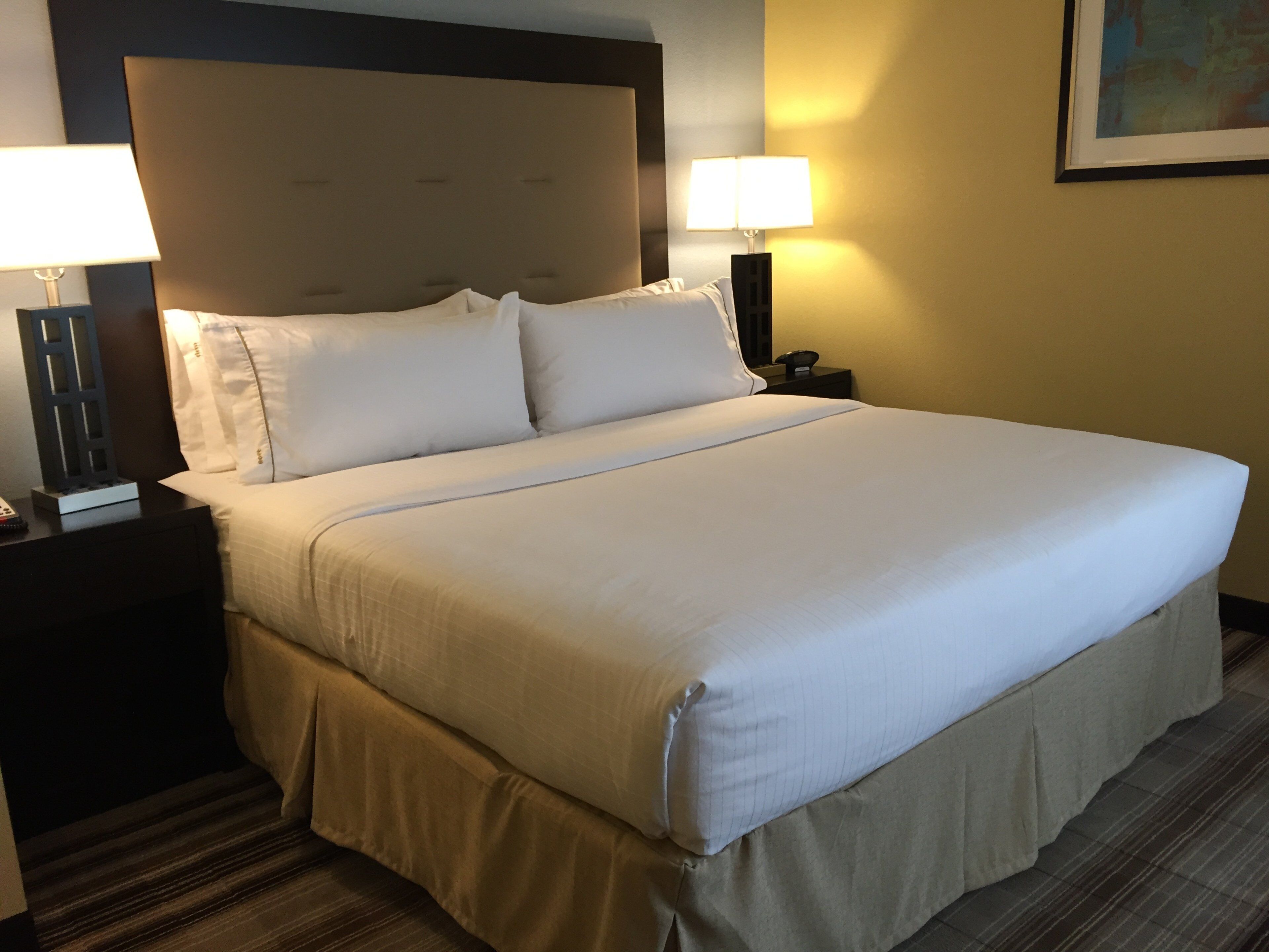 Quarto de hotel com uma cama grande, duas mesas de cabeceira, um painel de cabeceira e duas luminárias fixadas na parede.