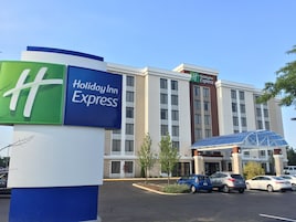 Edifício do Holiday Inn Express com céu claro, estacionamento com vários carros e uma grande placa com o logotipo do hotel.
