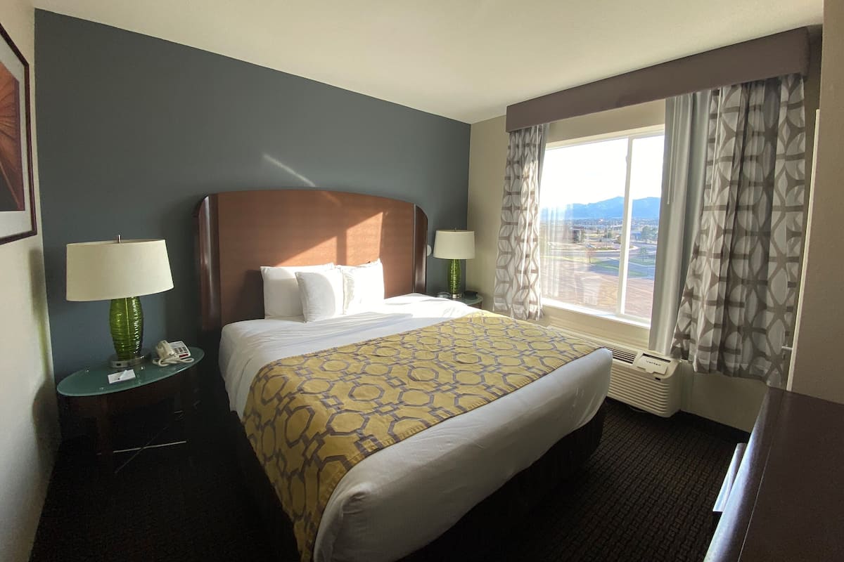 Quarto de hotel com uma cama grande, duas mesas de cabeceira com luminárias, uma janela com cortinas e vista para a paisagem urbana.