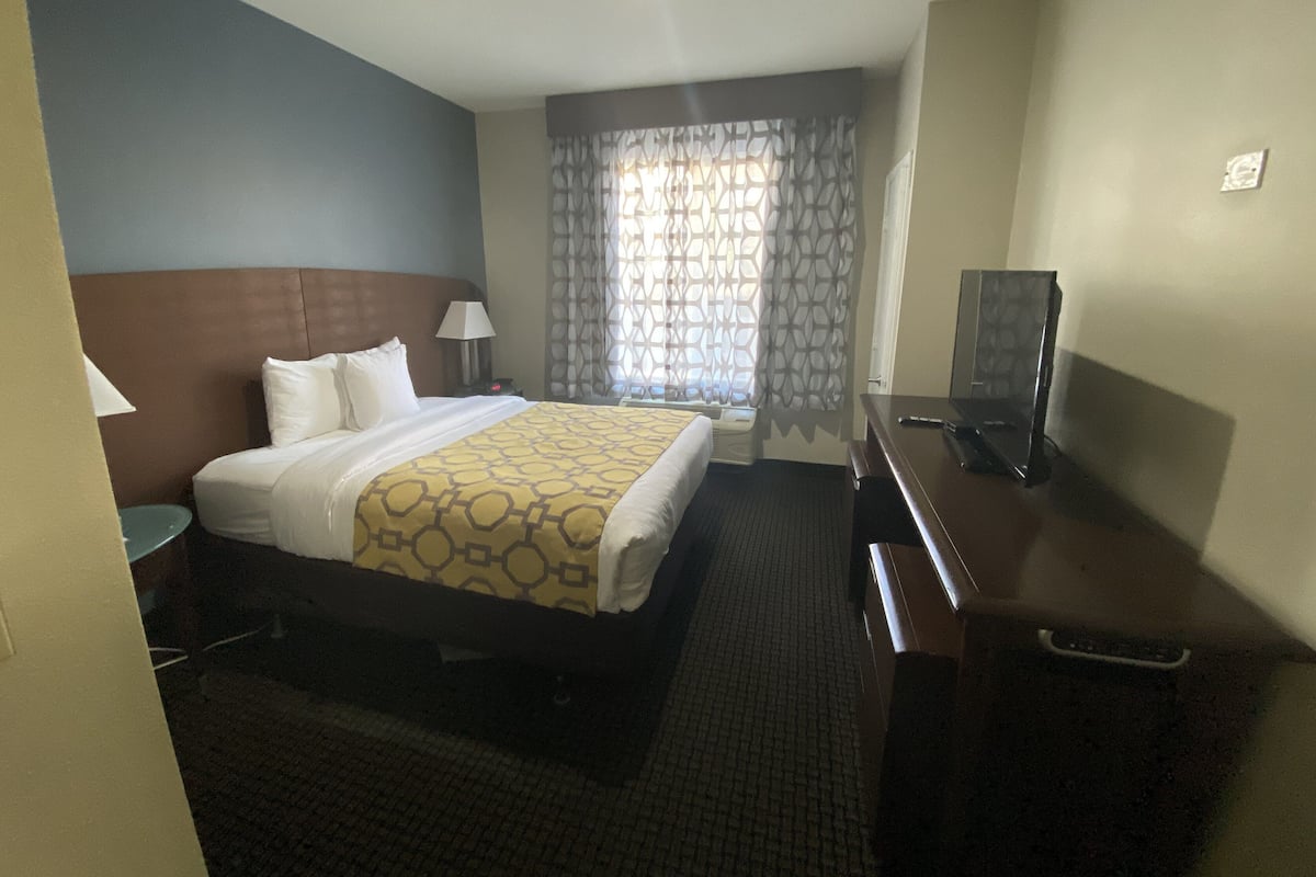 Quarto de hotel com uma cama grande, uma mesa de trabalho com televisão, uma luminária e uma janela com cortinas estampadas.