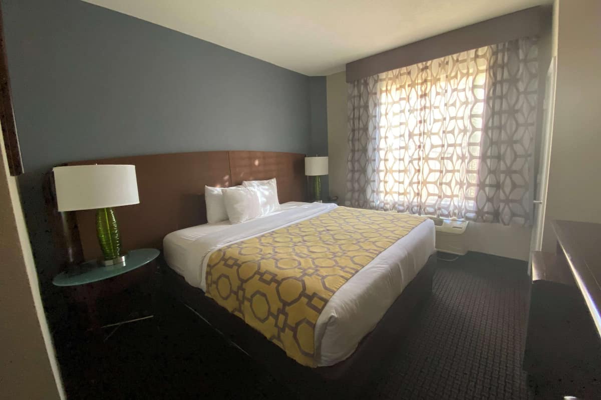 Quarto de hotel com cama, duas mesas de cabeceira, luminária, janela com cortinas estampadas e cabeceira.