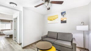 Uma sala de estar moderna com um sofá cinzento, um banquinho amarelo e um ventilador de teto. Há duas pinturas na parede e um abajur.