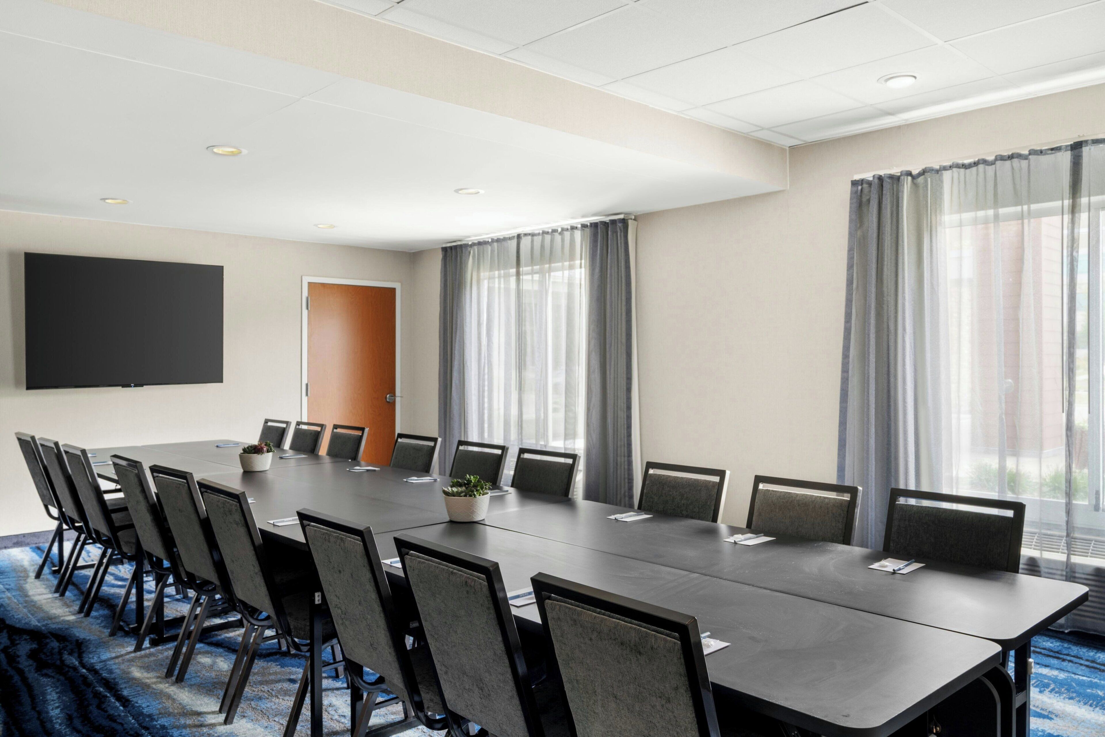 Uma sala de conferências com uma mesa comprida, cadeiras, uma TV de tela plana e grandes janelas com cortinas.