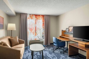 Quarto de hotel com sofá, mesa pequena, escrivaninha com cadeira, televisão e janela com cortinas.