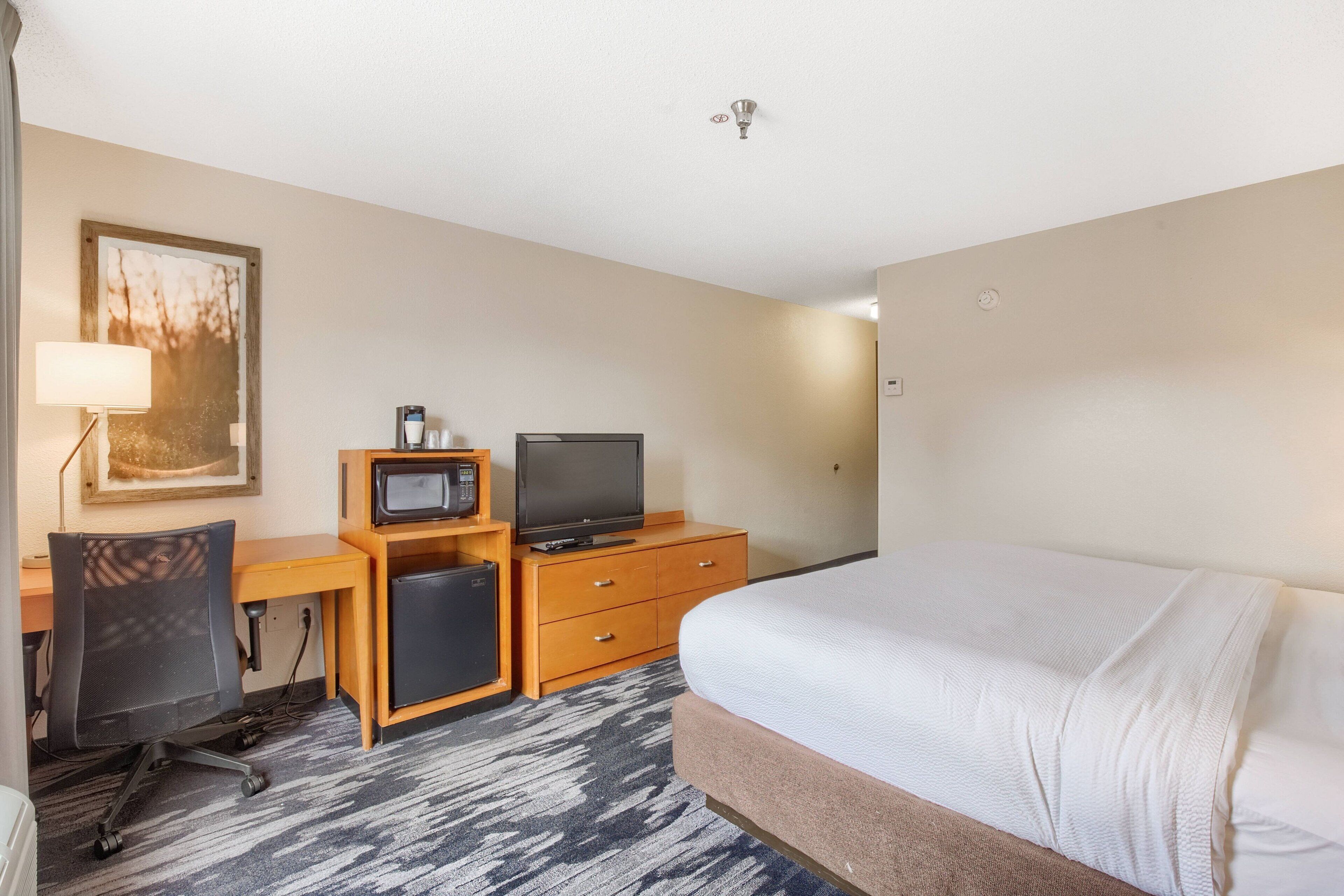 Quarto de hotel com uma cama, uma escrivaninha com computador, televisão e um pequeno frigorífico.