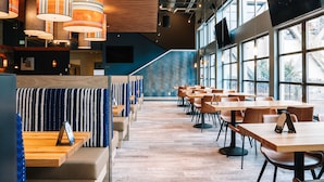 Interior de restaurante moderno com mesas de madeira, assentos listrados em azul e branco, e grandes janelas que permitem entrada de luz natural.