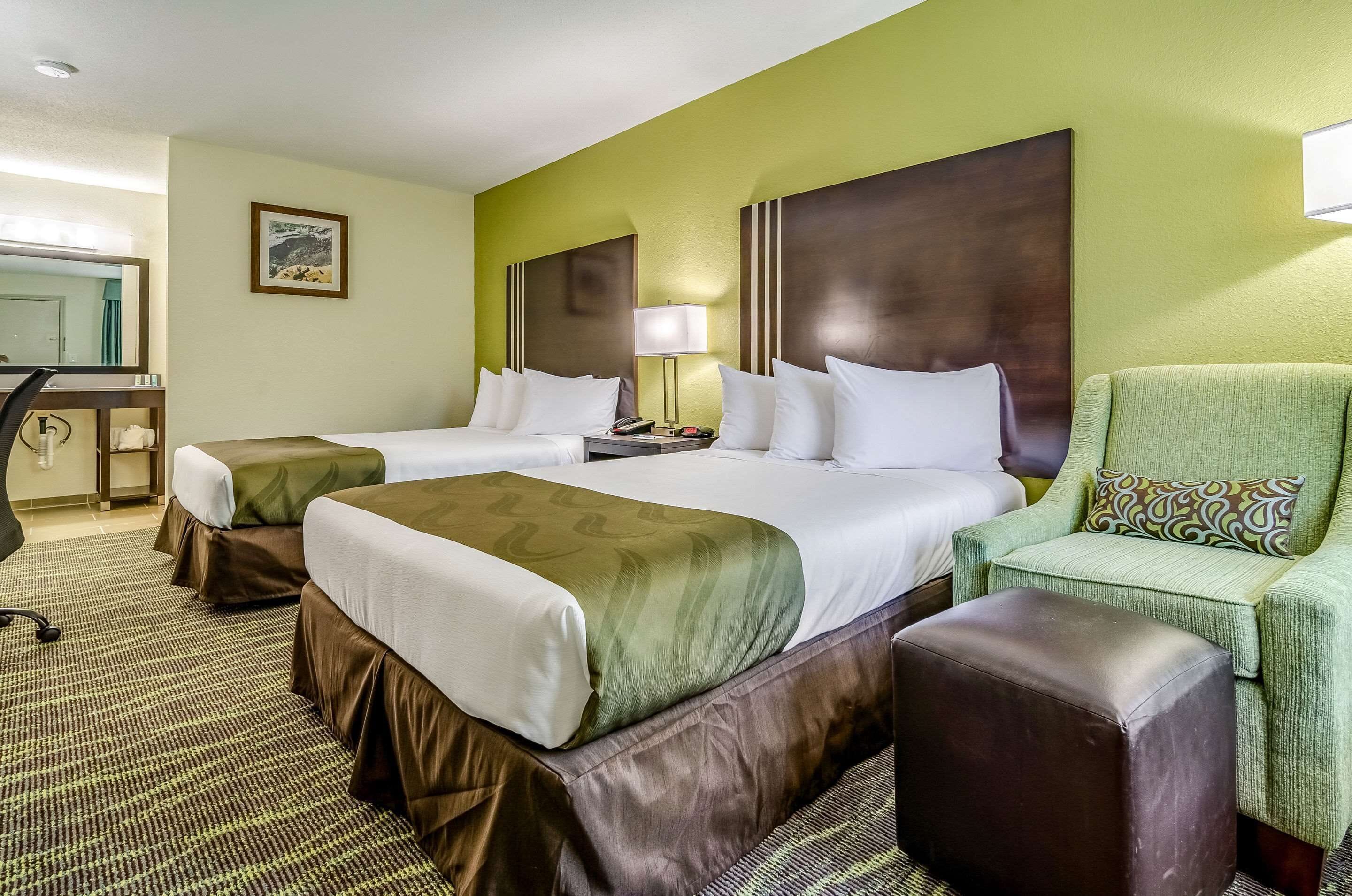 Quarto de hotel com duas camas, uma escrivaninha, uma cadeira e uma janela grande.