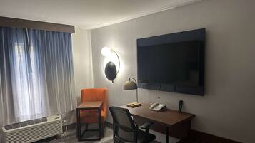 Quarto de hotel com escrivaninha, uma cadeira laranja, televisão fixada na parede, luminária e janela com cortinas.