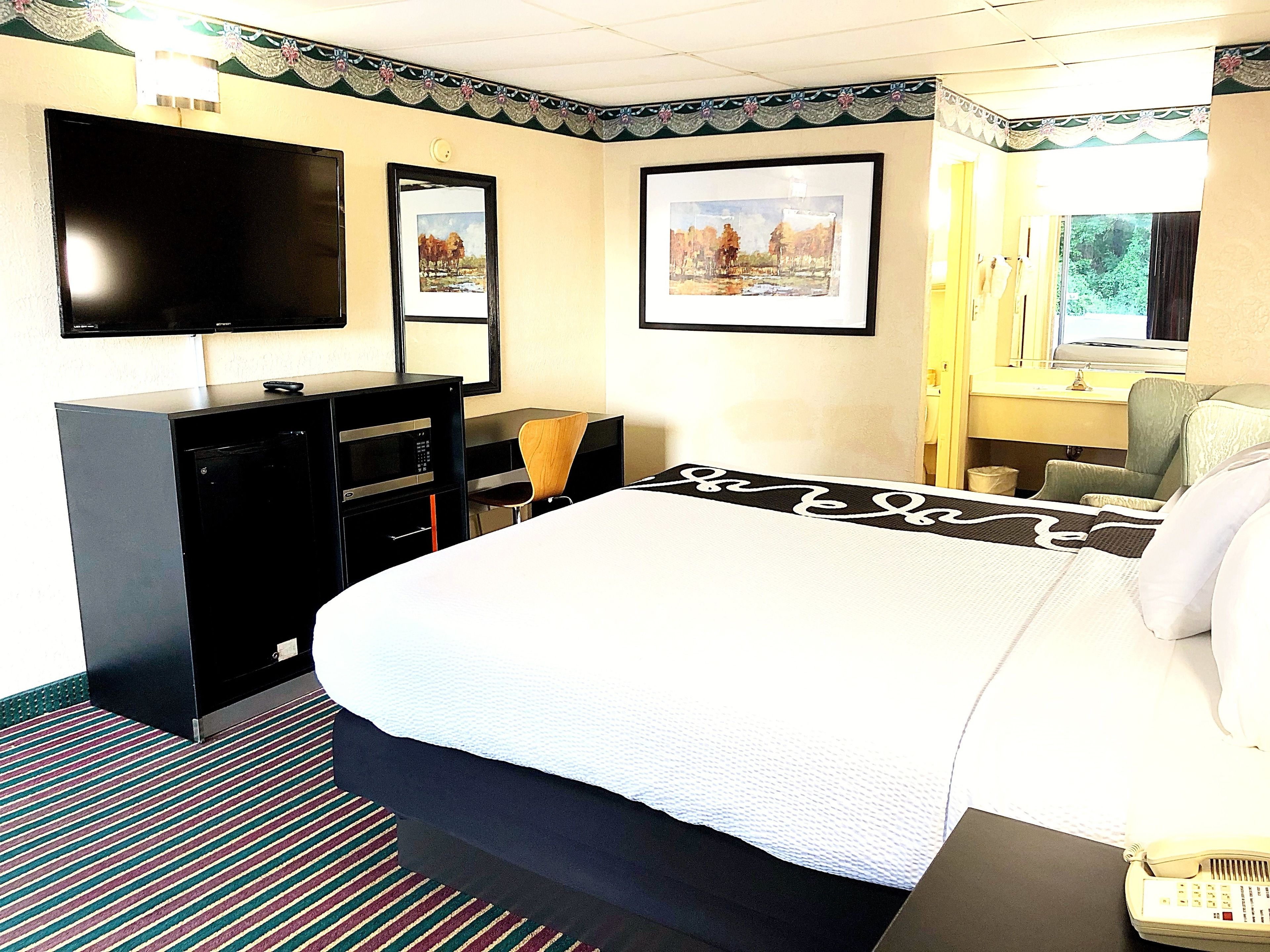 Quarto de hotel com uma cama grande, uma televisão de tela plana na parede, uma escrivaninha com cadeira e uma janela com vista para árvores.
