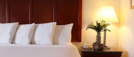 Cama bem arrumada com lençóis e almofadas brancas, cabeceira de madeira, mesa de cabeceira com luminária e uma pequena planta.