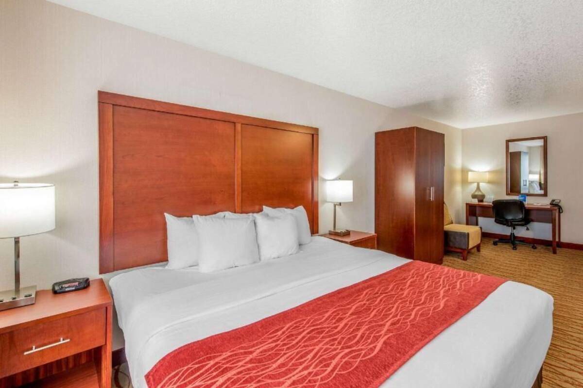 Quarto de hotel com uma cama grande, uma escrivaninha, uma cadeira, um guarda-roupa e um espelho.