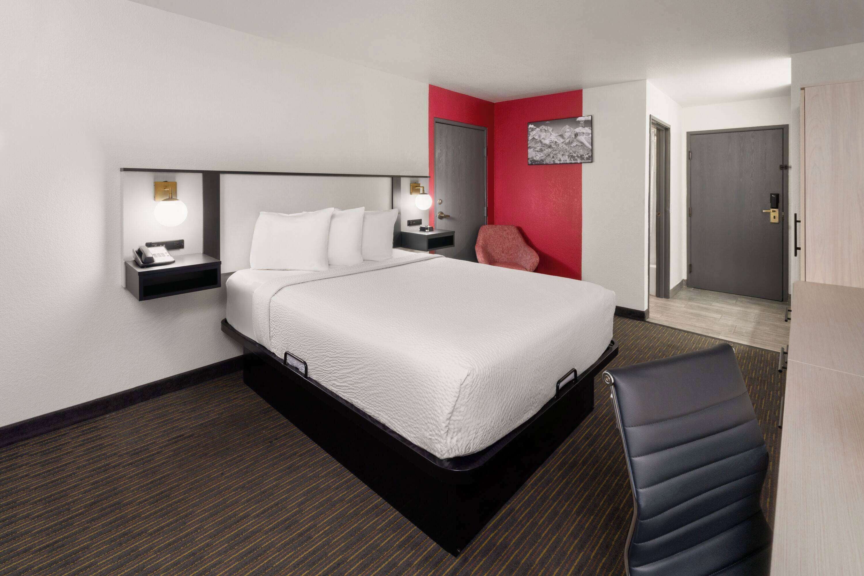 Quarto de hotel com uma cama grande, mesas de cabeceira com luminárias, uma parede de destaque vermelha, uma porta e uma cadeira.