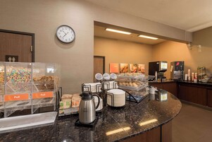 Espaço de café da manhã de hotel, com estação de café, cereais, doces e um relógio na parede.
