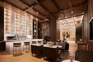 Uma área de bar moderna com bancada de mármore, banquetas e variedade de garrafas. Há áreas de estar com sofás e mesas, e uma grande tela de TV à direita.