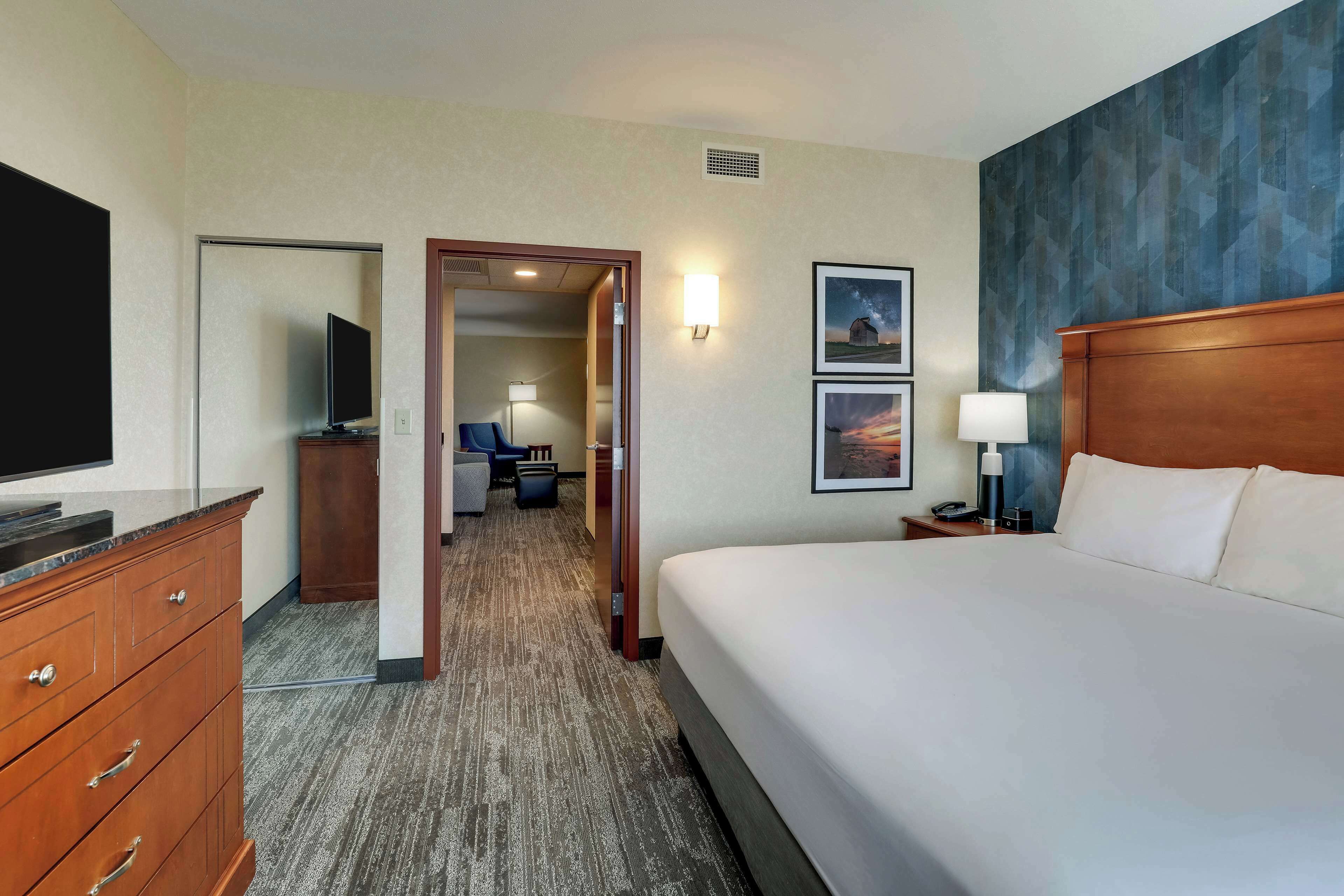Quarto de hotel com uma cama grande, televisão, escrivaninha com abajur e porta para um corredor.