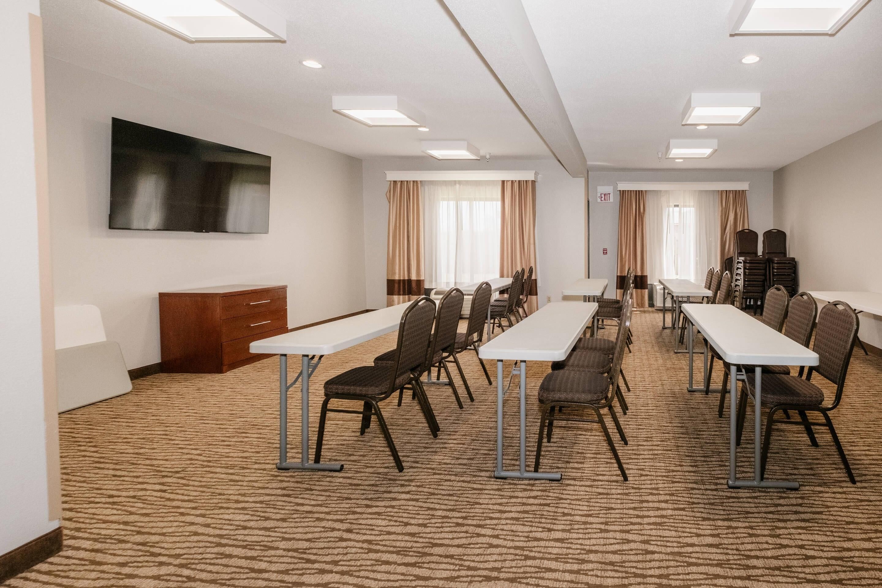 Uma sala de conferências com mesas compridas, cadeiras, uma TV de tela plana e um armário de madeira.