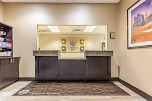 Uma área de recepção com a placa Comfort Inn, um balcão de atendimento, um quadro na parede e um tapete de boas-vindas.