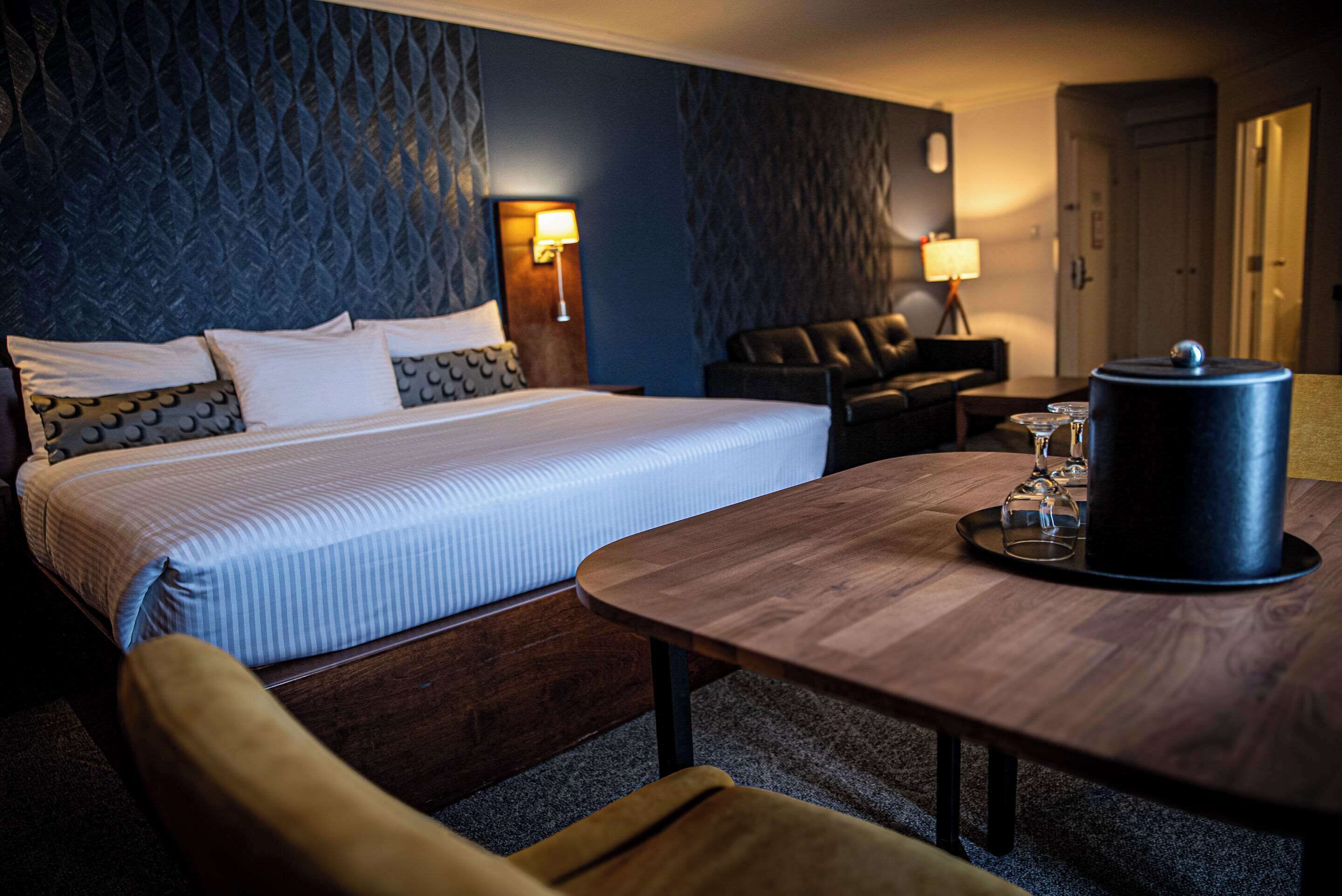 Quarto de hotel com uma cama grande, uma mesa de madeira com uma cafeteira, um sofá e duas luminárias.